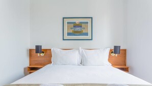 Cama bem arrumada com lençóis brancos, duas luminárias de apoio e uma obra de arte abstrata emoldurada na parede.