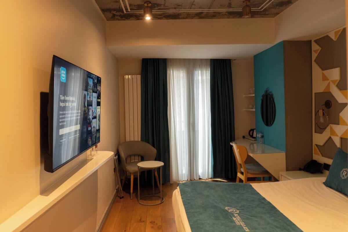 Quarto de hotel com cama, escrivaninha, cadeira, televisão e janela com cortinas.