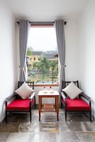 Quarto com duas cadeiras de madeira com almofadas vermelhas, uma pequena mesa com um vaso branco e uma flor vermelha, e vista de um edifício tradicional e palmeiras através de uma janela com cortinas cinzentas.