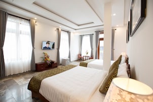 Quarto de hotel com duas camas, televisão, escrivaninha com cadeira e uma pequena mesa com um vaso de flores.