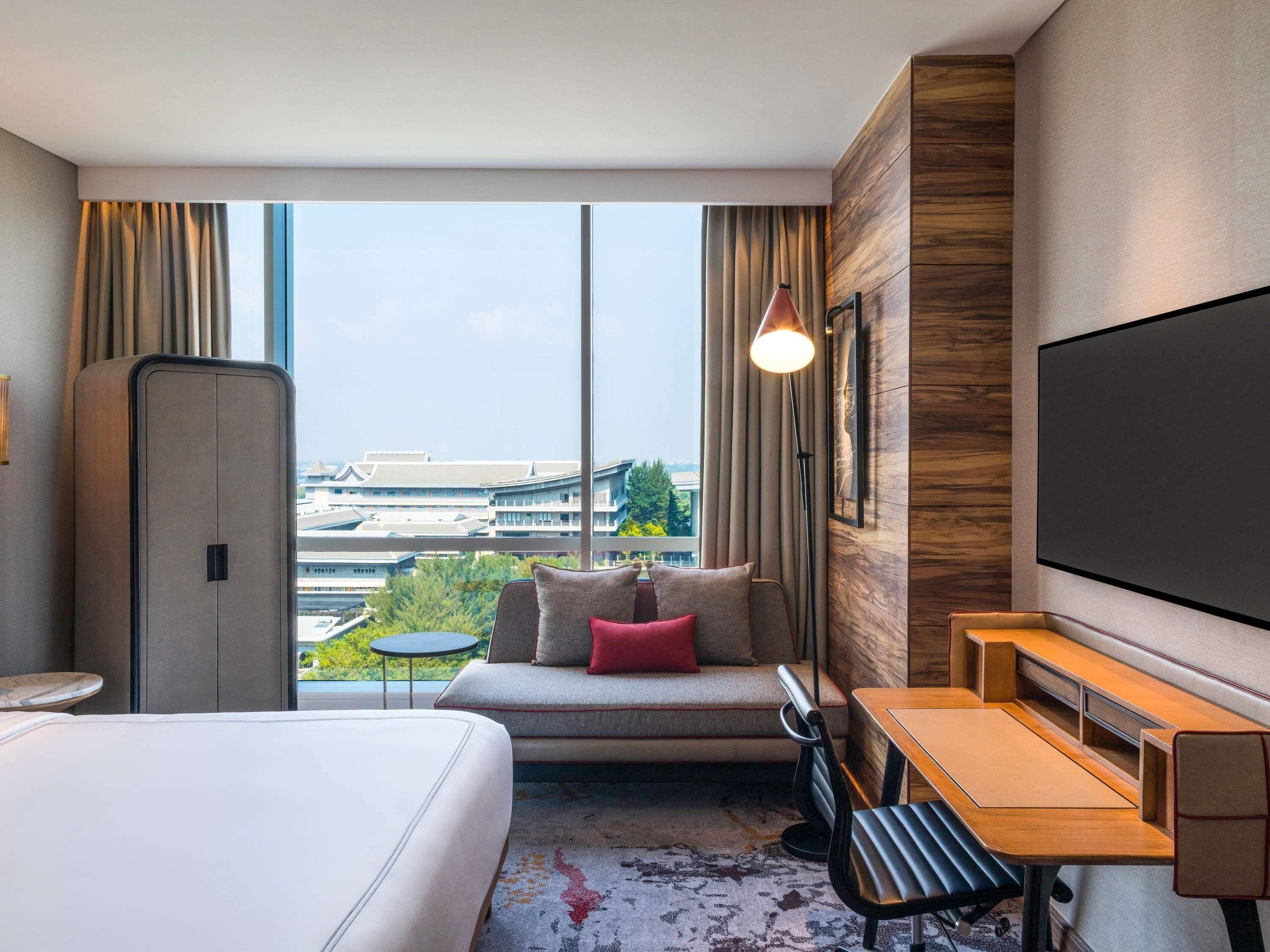 Quarto de hotel com cama, sofá, escrivaninha, TV e uma janela grande com vista para a cidade.