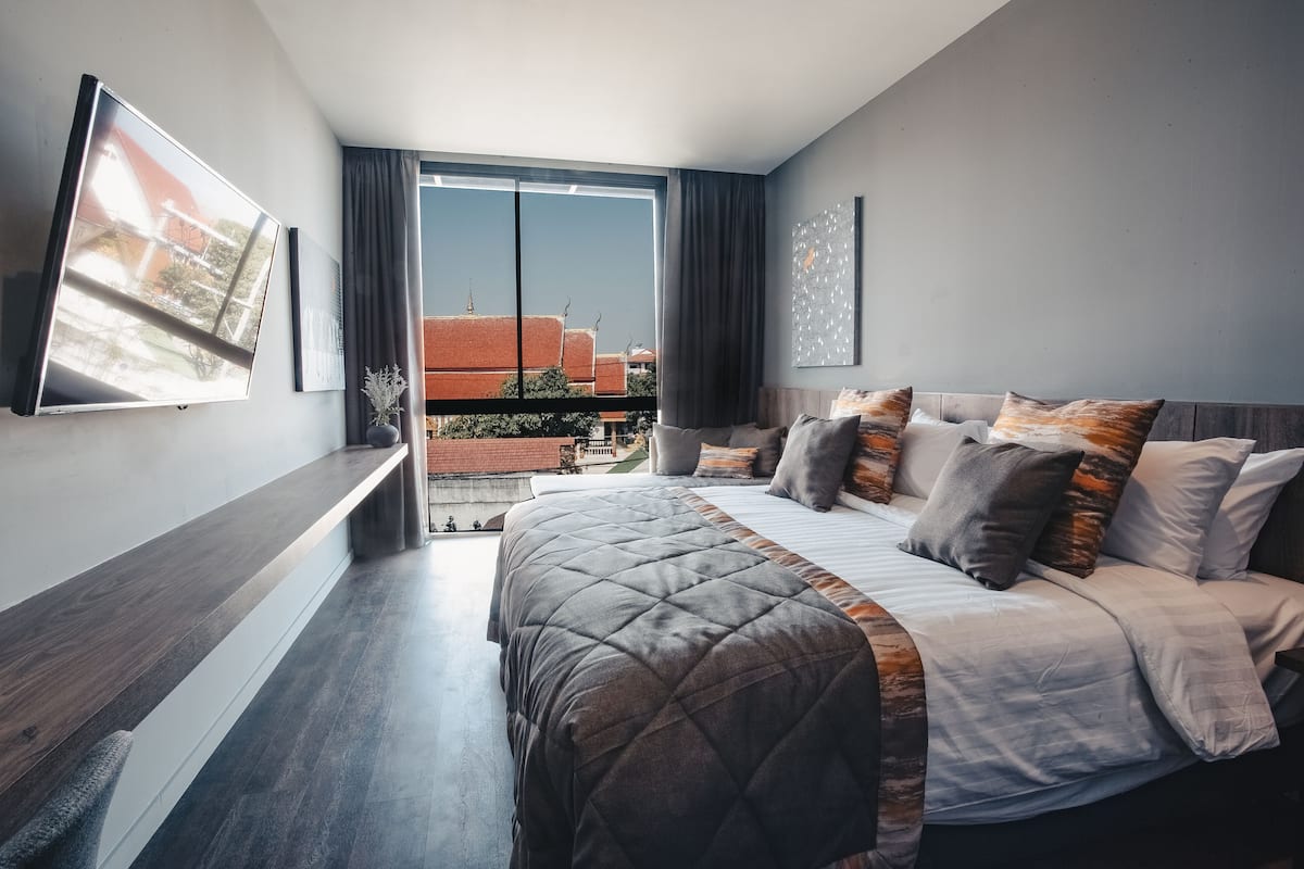 Quarto moderno com uma cama grande, uma TV na parede e vista para edifícios através de uma janela ampla.