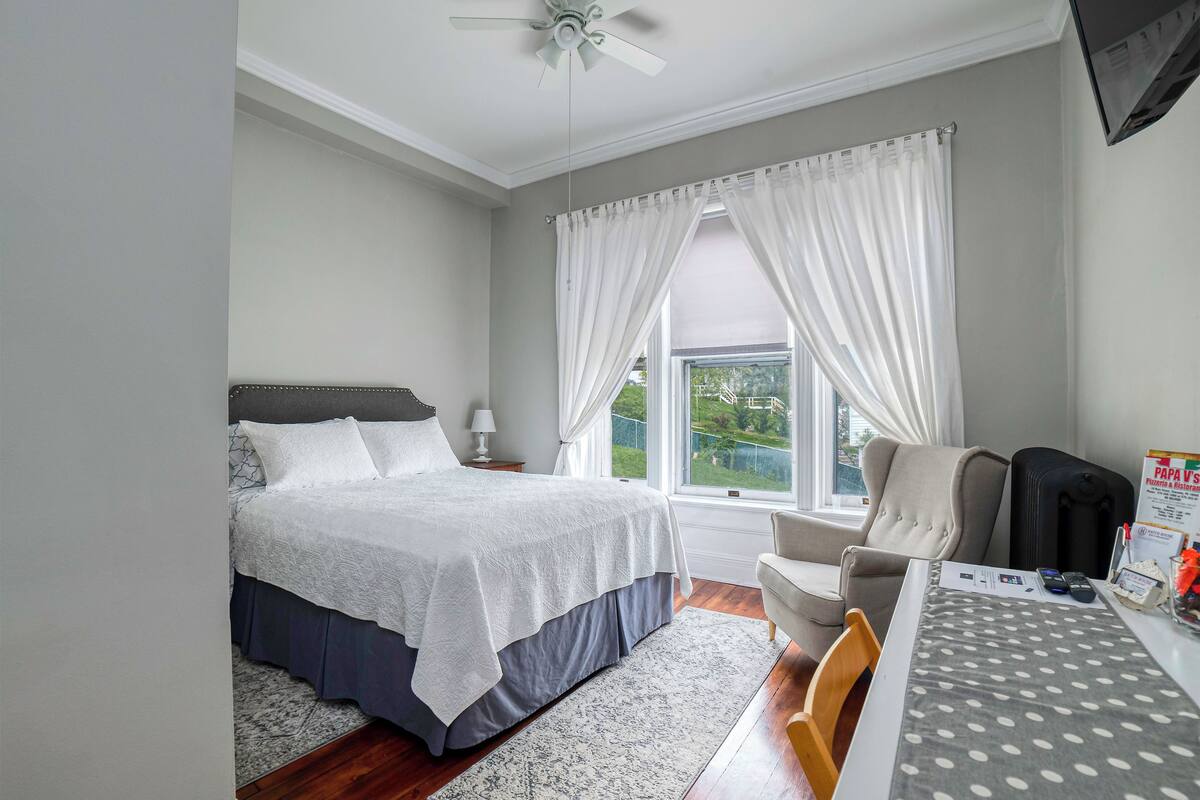 Quarto com cama, cadeira, escrivaninha e janela com cortinas.