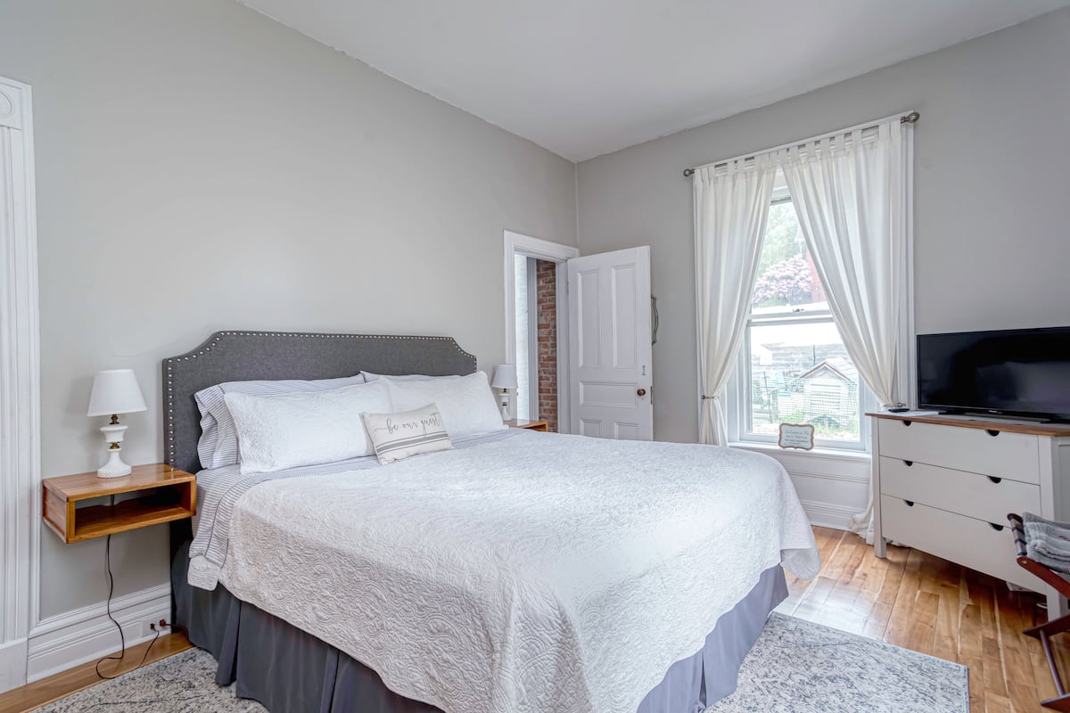 Quarto com cama, televisão, mesa de cabeceira, janela com cortinas e porta.