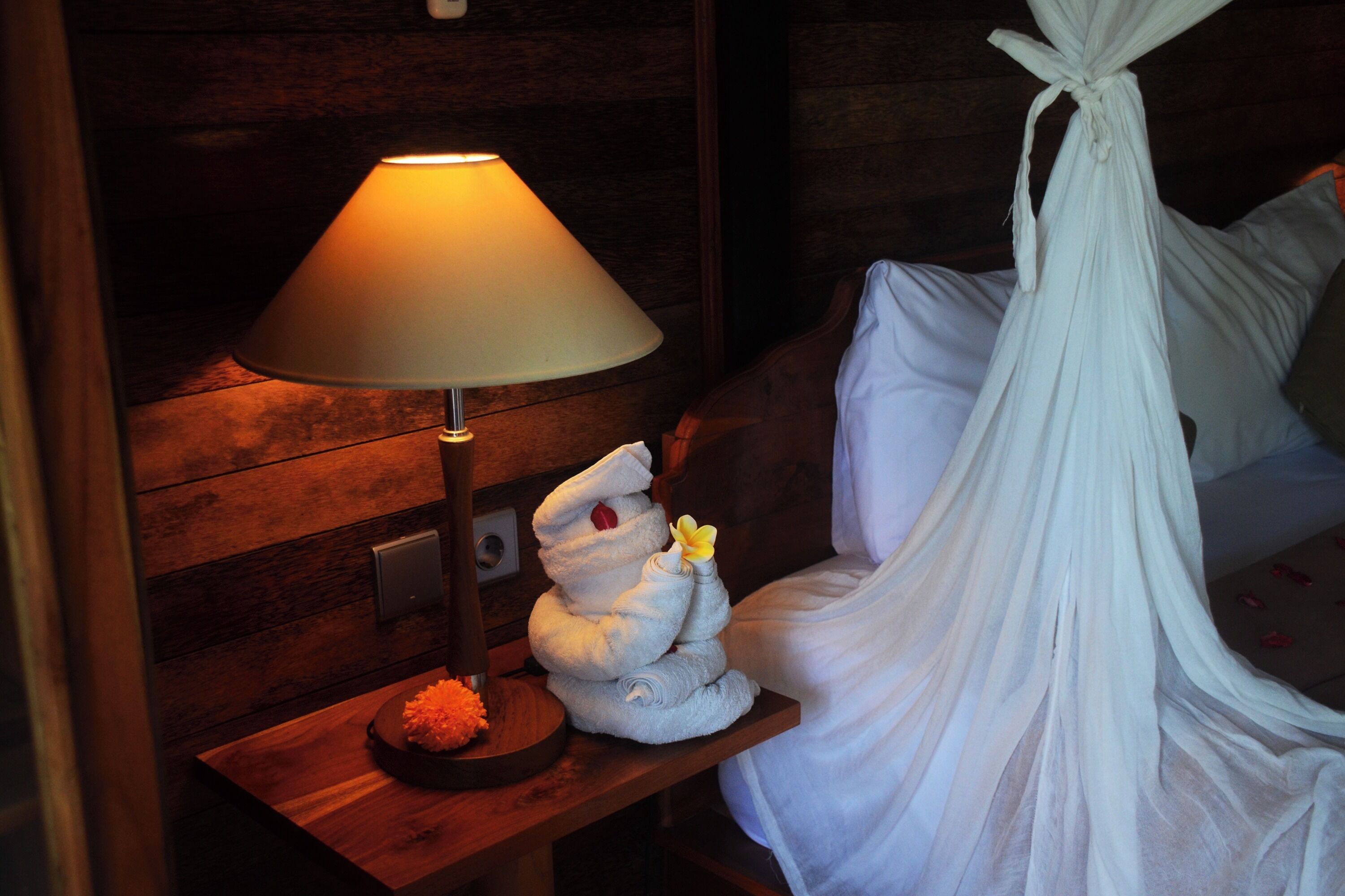 Uma parede de madeira, uma luminária de cabeceira, uma cama com dossel branco e uma escultura de toalha em um criado-mudo de madeira.