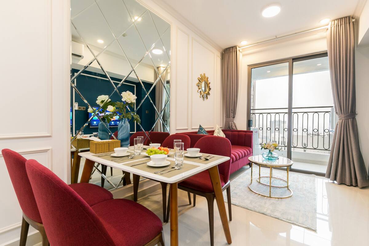 Uma área de refeições com uma mesa posta para quatro pessoas, cadeiras vermelhas estofadas, uma mesinha redonda com um vaso e vista para a varanda através de um painel de vidro.