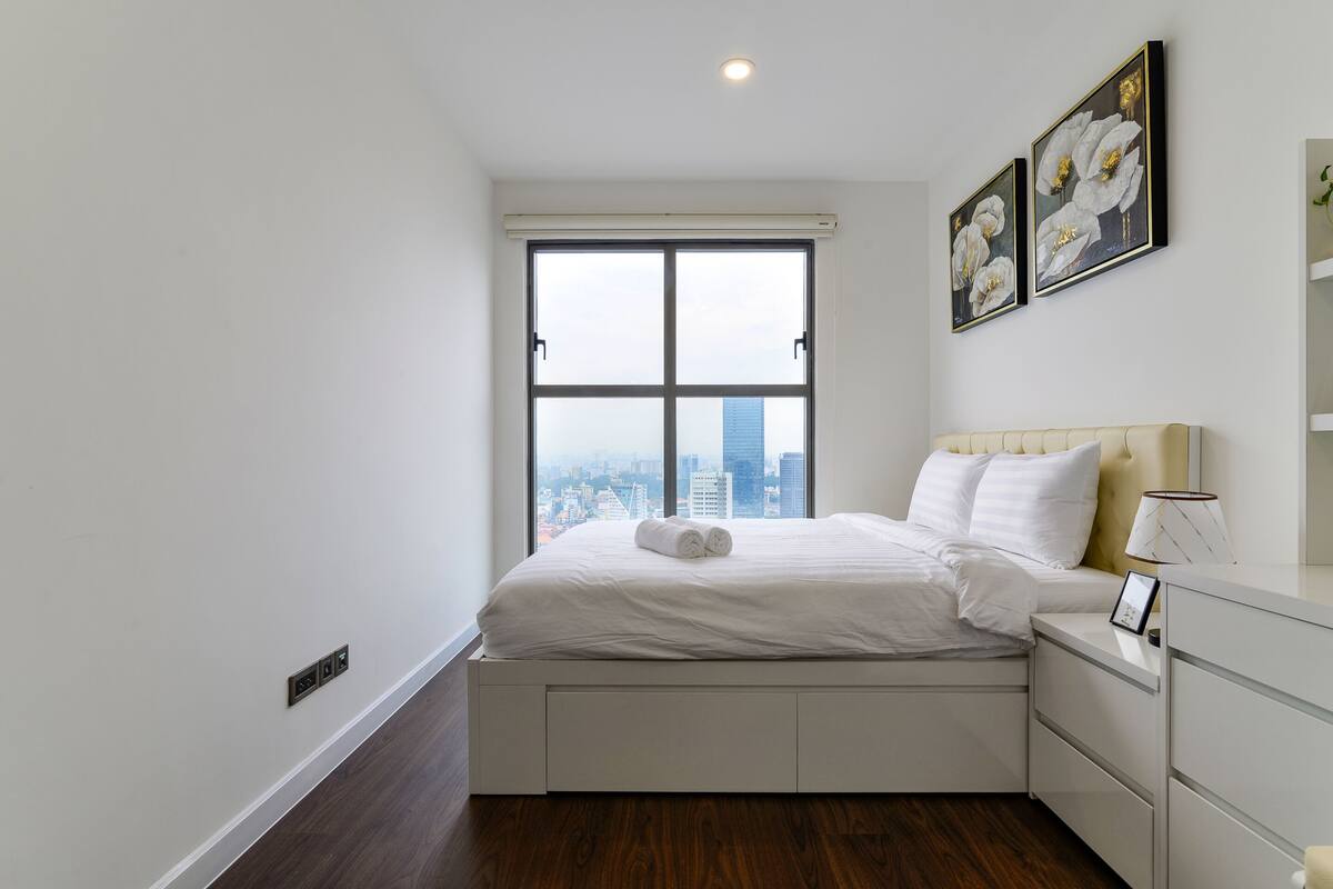 Quarto moderno com uma grande janela, cama com roupa de cama branca, mesa de cabeceira com abajur e duas obras de arte florais emolduradas na parede.
