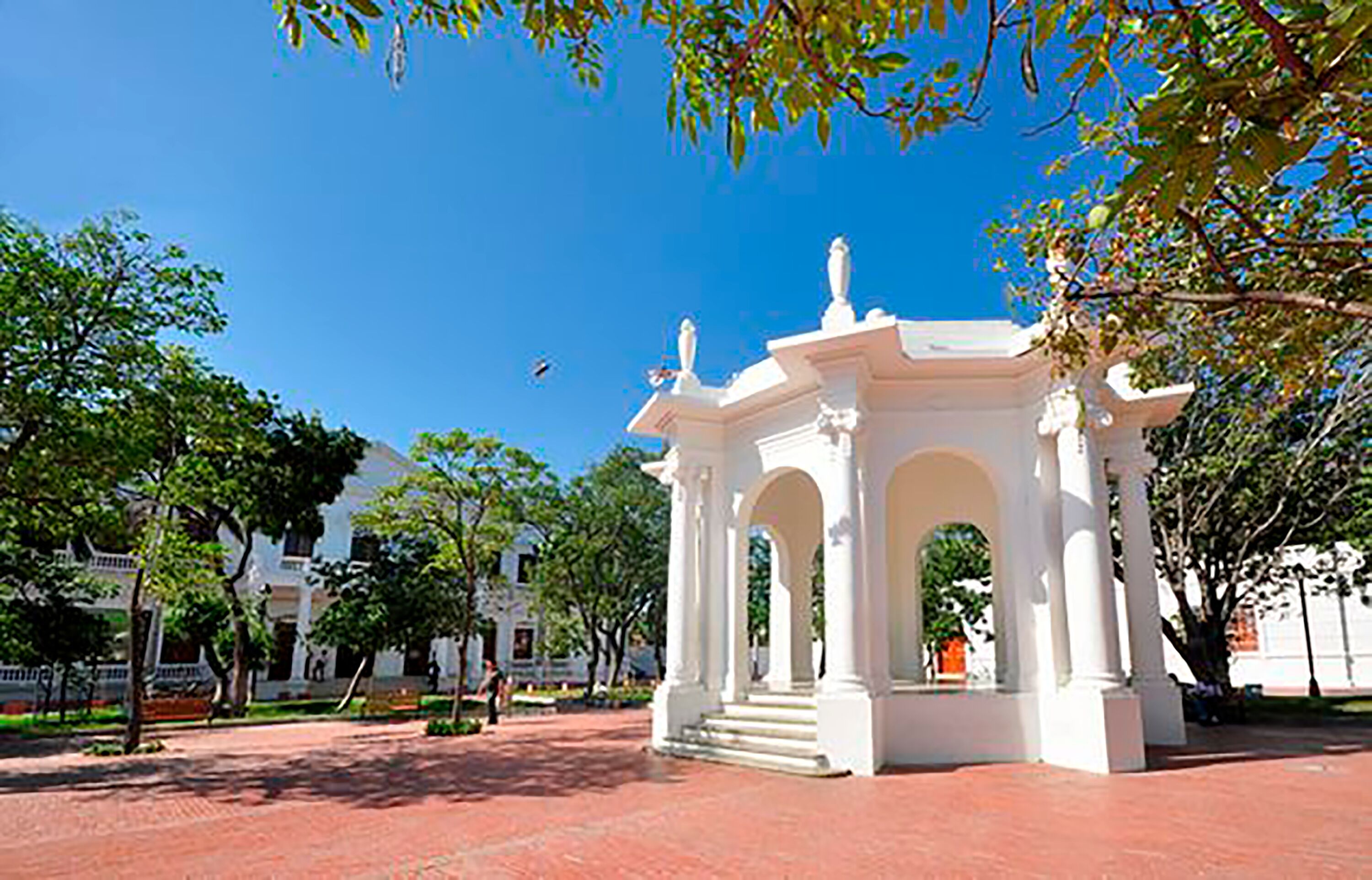 Um gazebo branco com colunas e uma estátua no topo, rodeado por árvores e um caminho de tijolos.
