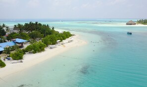 Um resort à beira-mar com areal arenoso, águas turquesa cristalinas e algumas embarcações. Há construções com telhados azuis e guarda-sóis ao longo da praia.