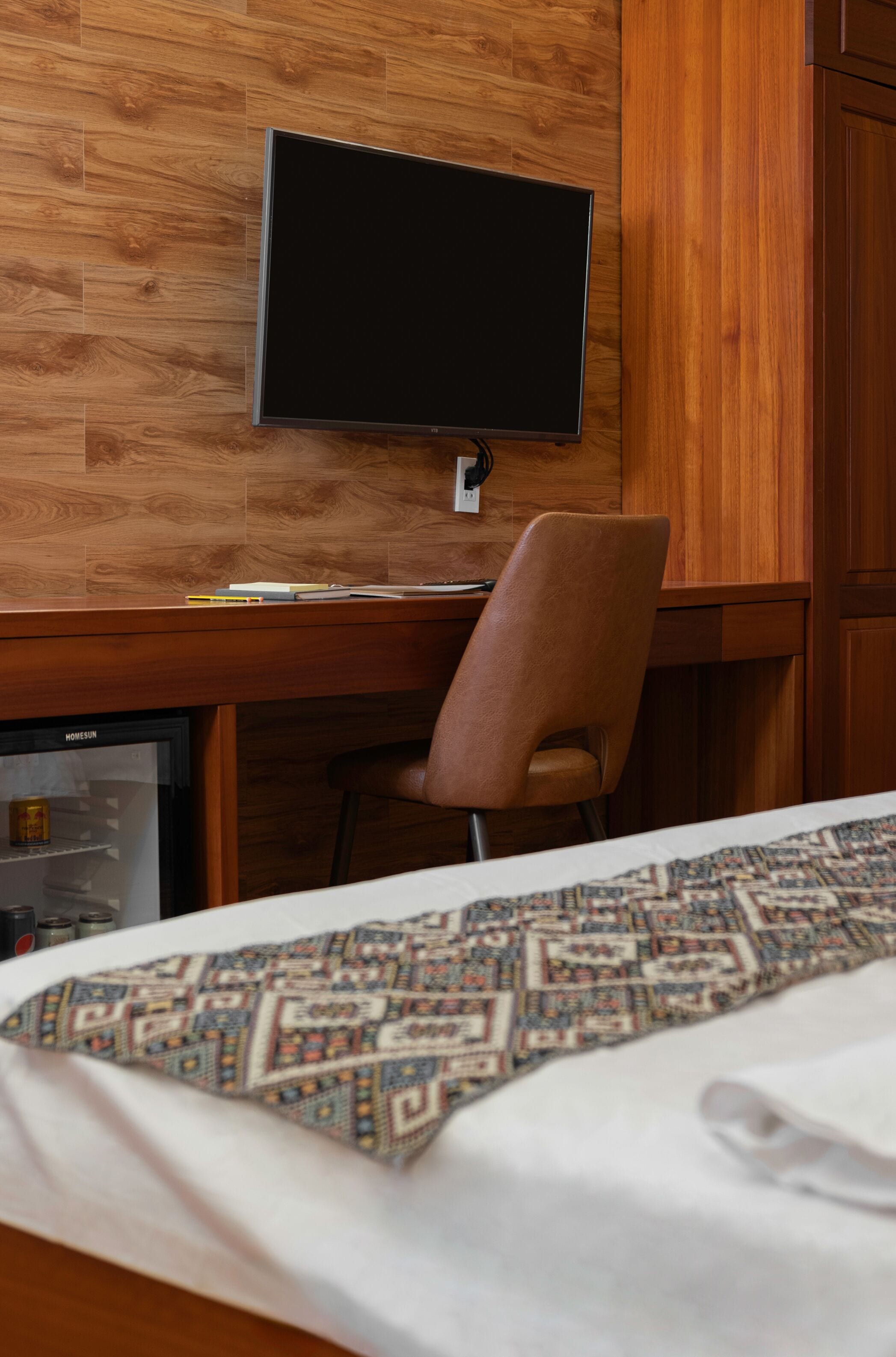 Quarto de hotel com cama, escrivaninha com cadeira, televisão na parede e minibar.