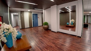 Um lobby moderno com piso de madeira, um balcão de recepção com vasos de flores, uma grande planta em vaso e um espelho refletindo o espaço.