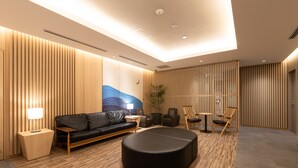 Quarto de hotel moderno com sofá de couro preto, um banquinho redondo e piso de madeira. Há duas mesinhas com abajures, um quadro grande na parede e uma porta deslizante ao fundo.