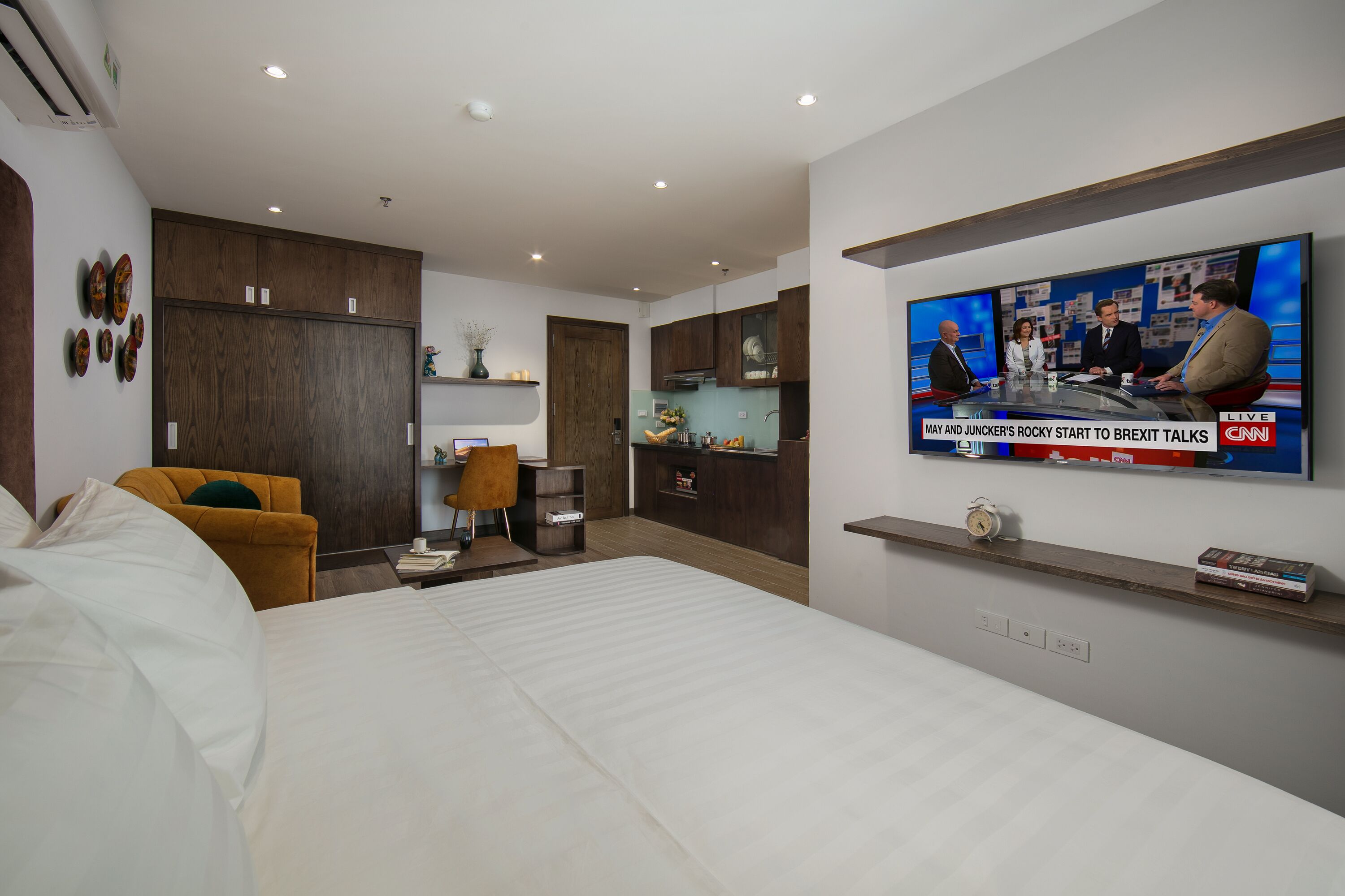 Quarto de hotel moderno com uma cama grande, televisão fixada na parede, mobiliário de madeira e uma pequena escrivaninha com cadeira.