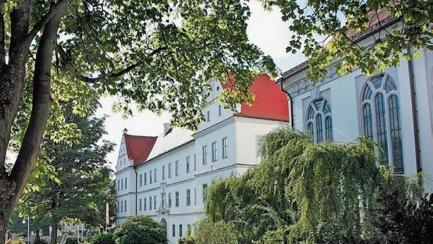 Um edifício grande e histórico com telhado vermelho e paredes brancas, rodeado por vegetação exuberante e um caminho de acesso pavimentado.