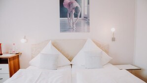 Quarto de hotel com duas camas, uma mesa de cabeceira de madeira, uma mesinha e uma luminária na parede.