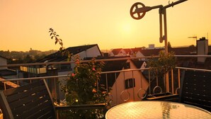 Terraço no topo com mesa de vidro redonda, plantas em vasos e vista do horizonte ao pôr do sol.