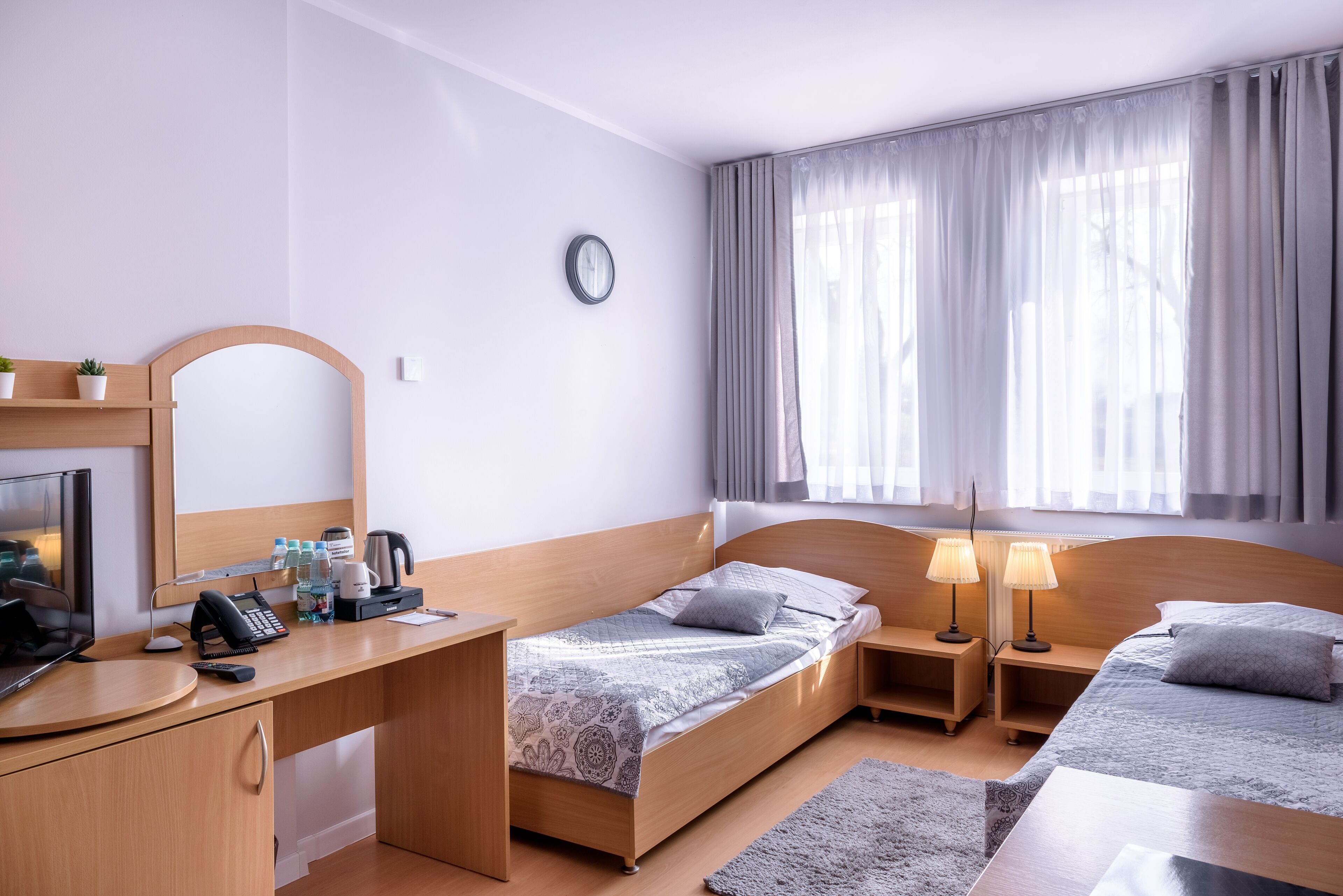 Quarto de hotel com escrivaninha, duas camas de solteiro, um pequeno espaço de estar, uma janela grande com cortinas e um relógio na parede.