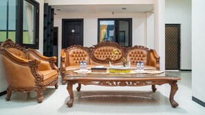 Um quarto com um sofá grande e ricamente decorado, poltronas combinando, uma mesa de centro de vidro com objetos decorativos e uma porta que dá para outro cômodo.