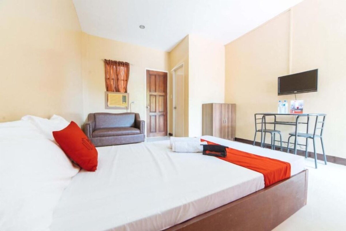 Quarto de hotel com uma cama grande, uma escrivaninha com cadeiras, uma televisão e um guarda-roupa.