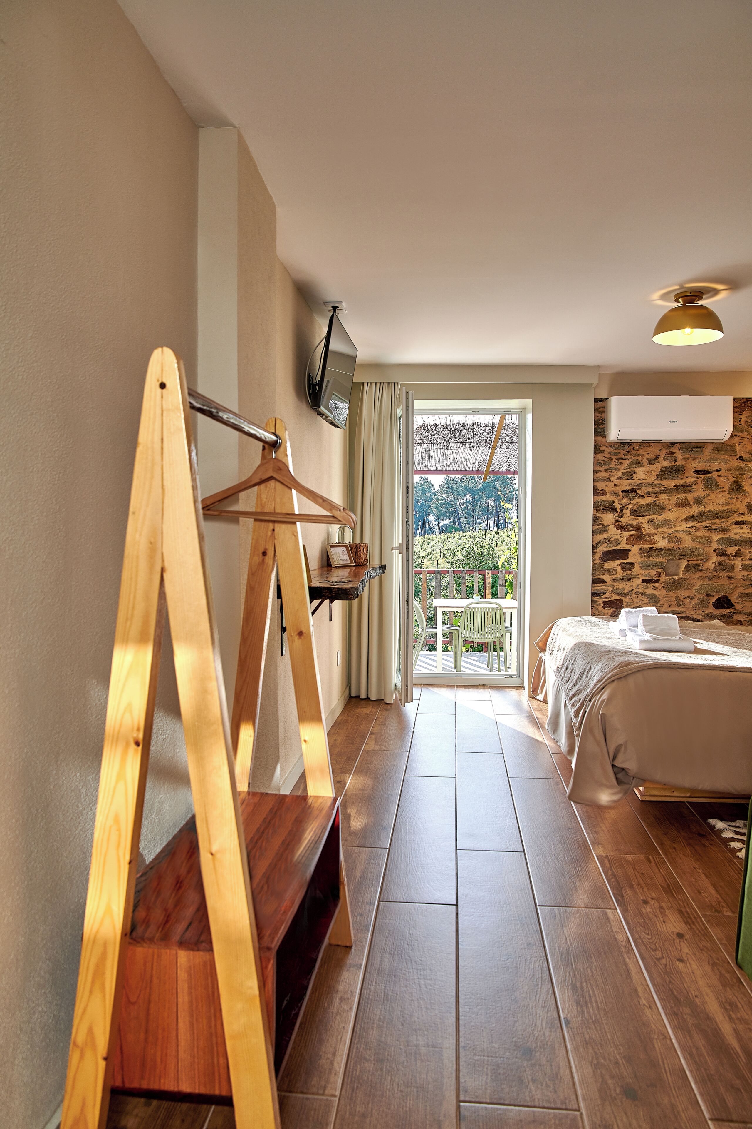 Quarto com cama, um cabide de madeira, mesa com cadeira e vista para o verde lá fora.