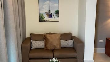 Uma sala de estar com um sofá marrom, uma mesa de centro de vidro com um vaso, um quadro de um veleiro e um lustre.