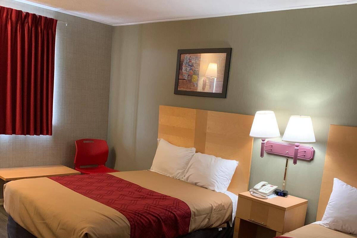 Quarto de hotel com duas camas, uma cadeira vermelha, mesa de cabeceira com abajur e detector de fumaça no teto.