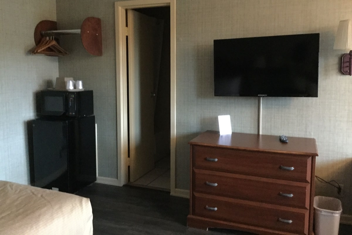 Quarto de hotel com cama, cómoda de madeira, televisão, micro-ondas e porta que dá para outro espaço.