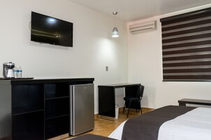 Quarto de hotel com cama, escrivaninha, televisão, frigorífico e janela com persianas.