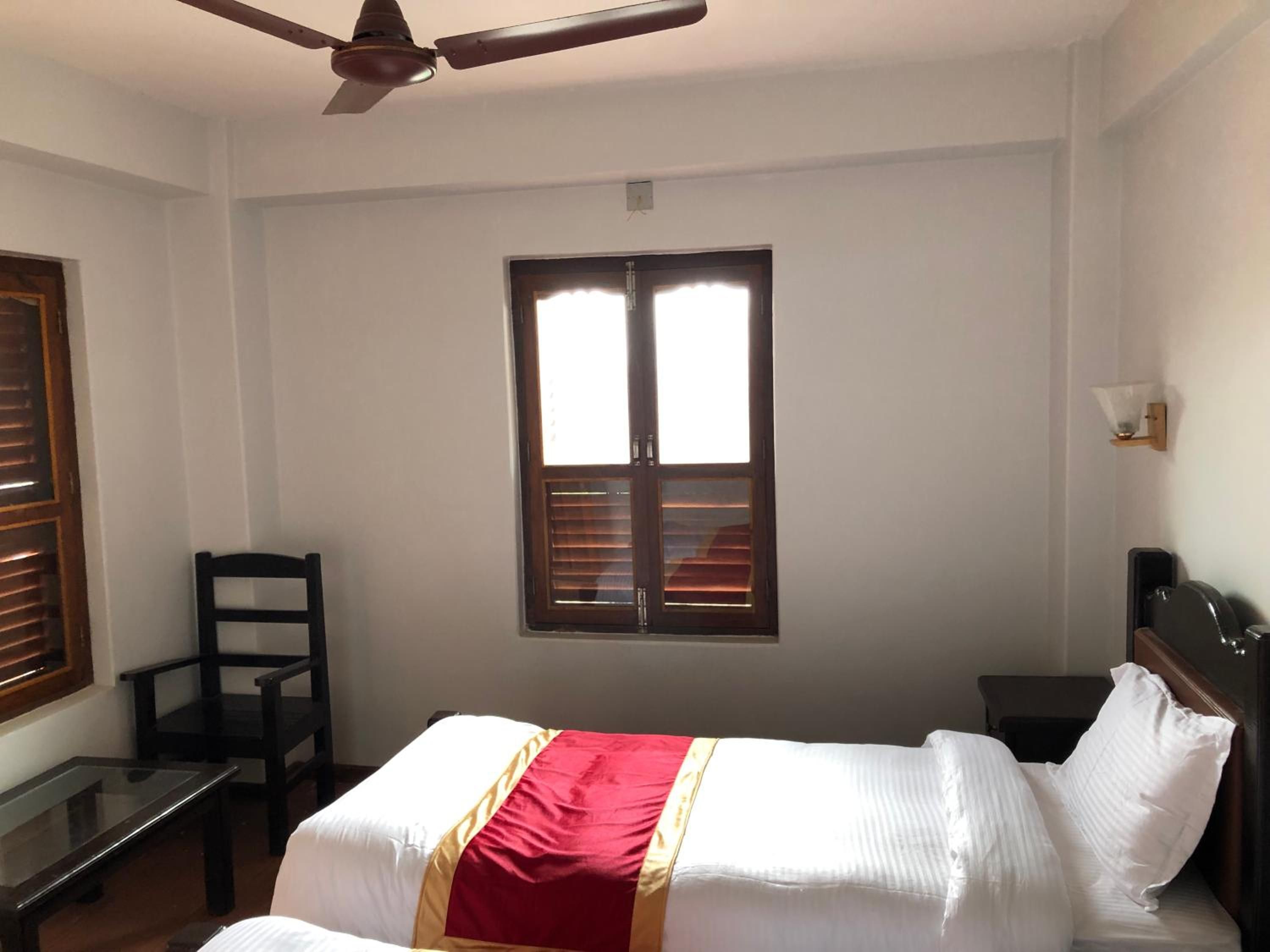 Quarto com cama, cadeira, mesa de cabeceira, ventilador de teto e janela com persianas.
