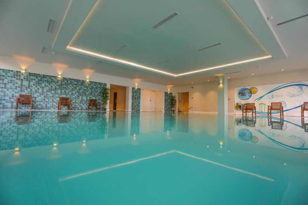 Uma grande piscina interna com água cristalina, rodeada por cadeiras de madeira e uma parede revestida com um padrão de ondas.