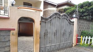 Uma entrada gradeada com portão de metal e um caminho de paralelepípedos que leva a uma casa com fachada de pedra e varanda.