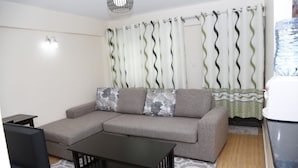 Uma sala de estar com um sofá modular cinza, uma mesa de centro preta, uma televisão e um tapete estampado.