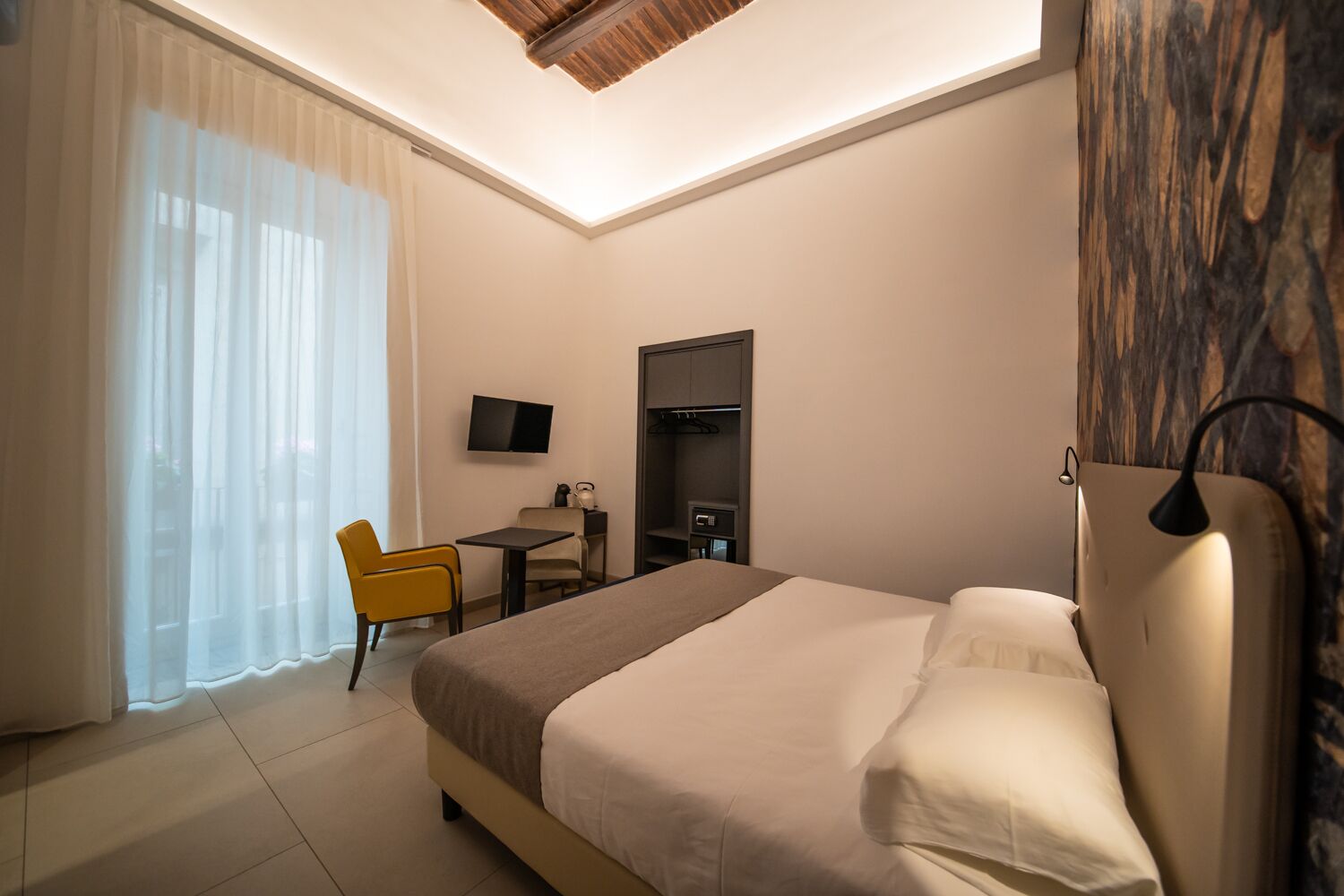 Quarto de hotel com cama, escrivaninha, cadeira, televisão e guarda-roupa.