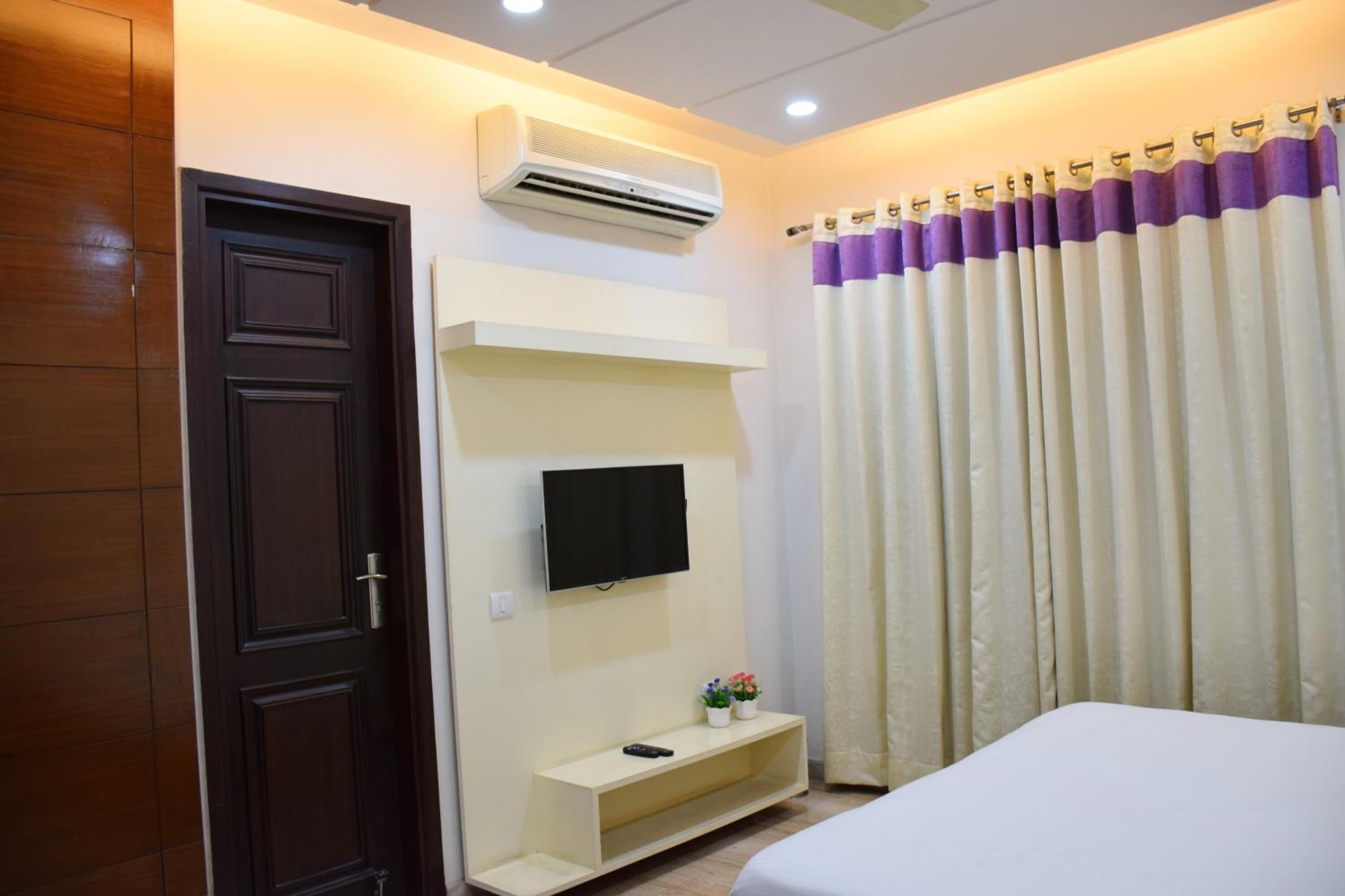 Quarto de hotel com porta de madeira escura, ar condicionado fixado na parede, televisão de tela plana em parede branca, prateleira pequena com controle remoto e flores, e cama com colchão branco.