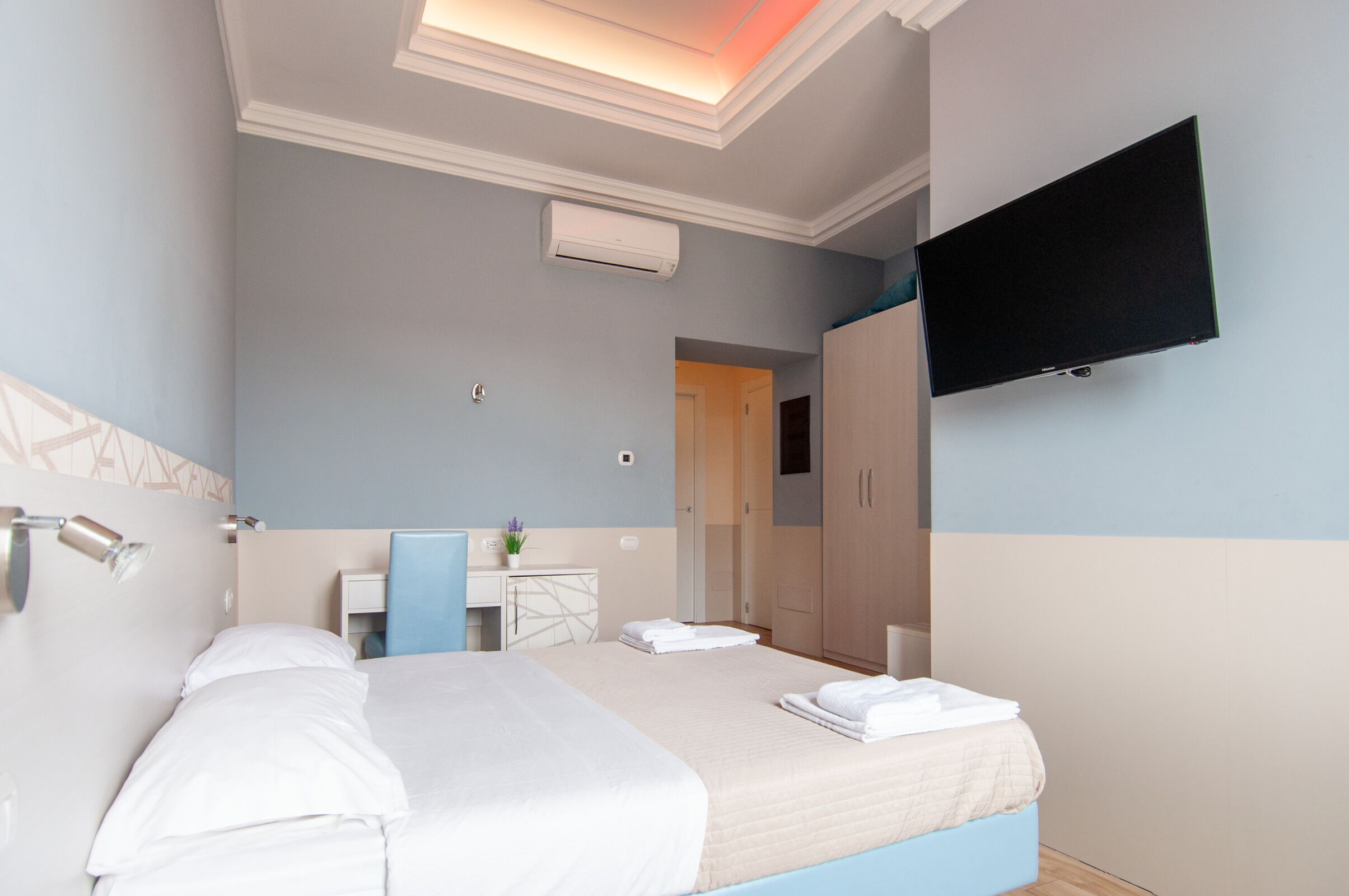 Quarto de hotel com cama, televisão de tela plana na parede, escrivaninha com cadeira e guarda-roupa.