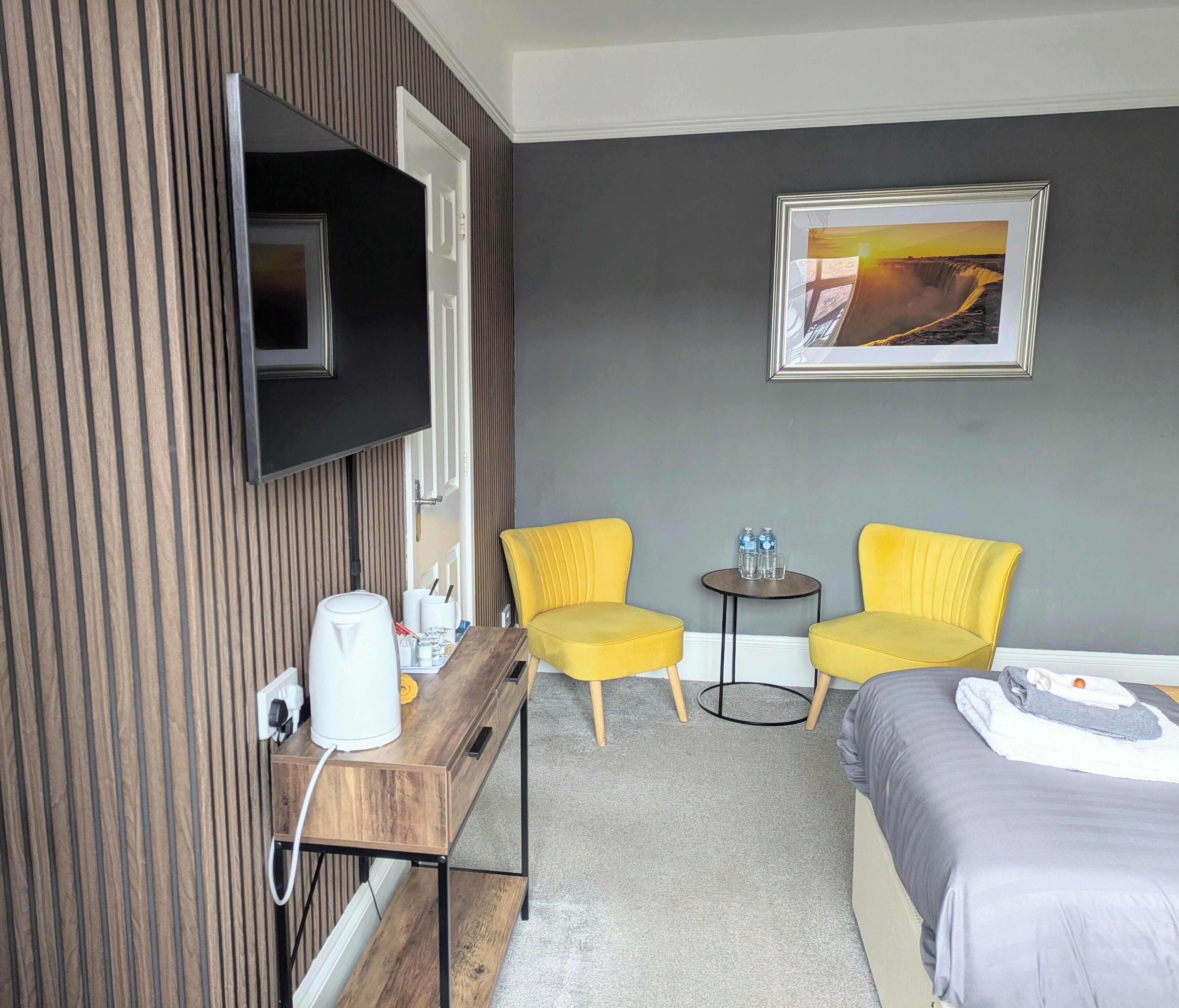 Quarto de hotel com cama, escrivaninha com cafeteira, duas poltronas amarelas, mesa redonda pequena com garrafas de água e um quadro na parede.