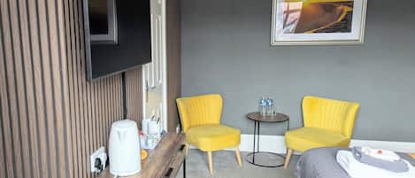 Quarto de hotel com cama, escrivaninha com cafeteira, duas poltronas amarelas, mesa redonda pequena com garrafas de água e um quadro na parede.