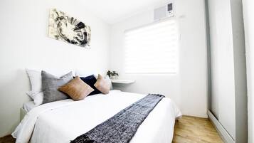 Quarto com cama, almofadas, cobertor, uma obra de arte na parede, janela com persianas e uma pequena mesa de cabeceira.
