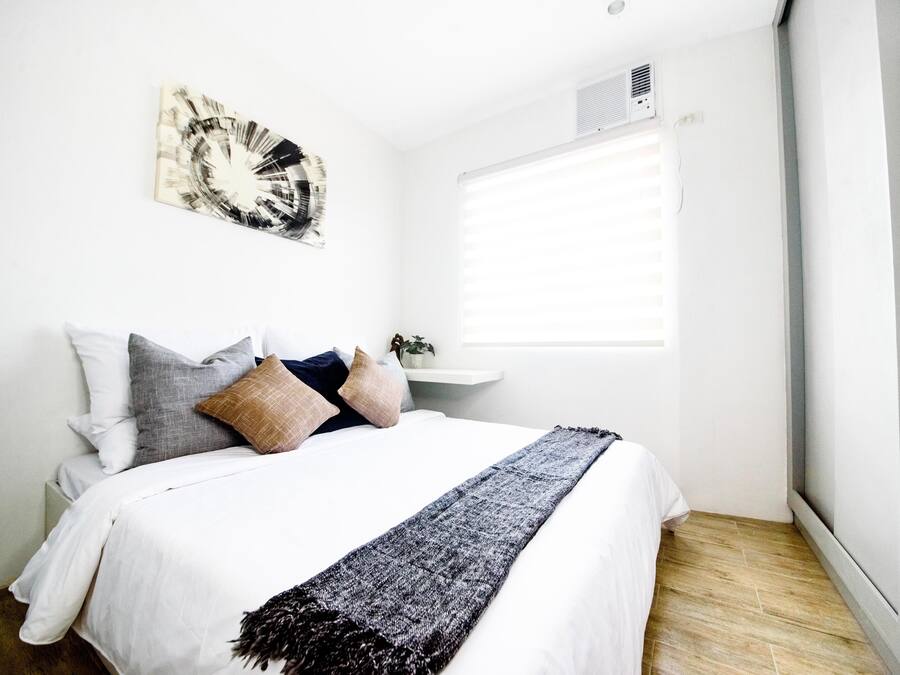 Quarto com cama, almofadas, cobertor, uma obra de arte na parede, janela com persianas e uma pequena mesa de cabeceira.