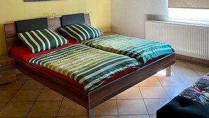 Quarto com cama, mesa de cabeceira, janela, radiador e ventoinha no teto.