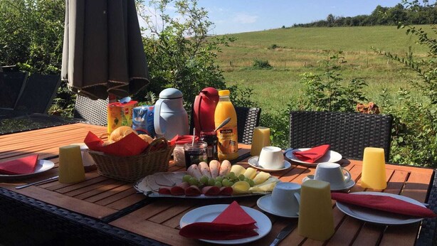 Mesa de refeições ao ar livre posta com comida e bebidas, rodeada por cadeiras de vime e um grande guarda-sol.