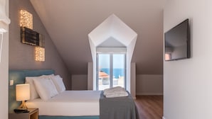 Quarto com cama, mesa de cabeceira, televisão e vista para o mar através de uma janela.