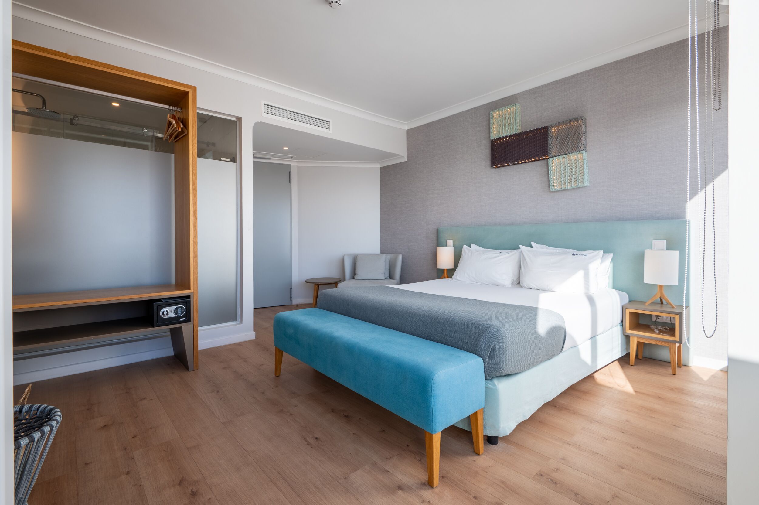 Quarto de hotel moderno com uma cama grande, um banco, um closet espaçoso e uma mesa de cabeceira com luminária.