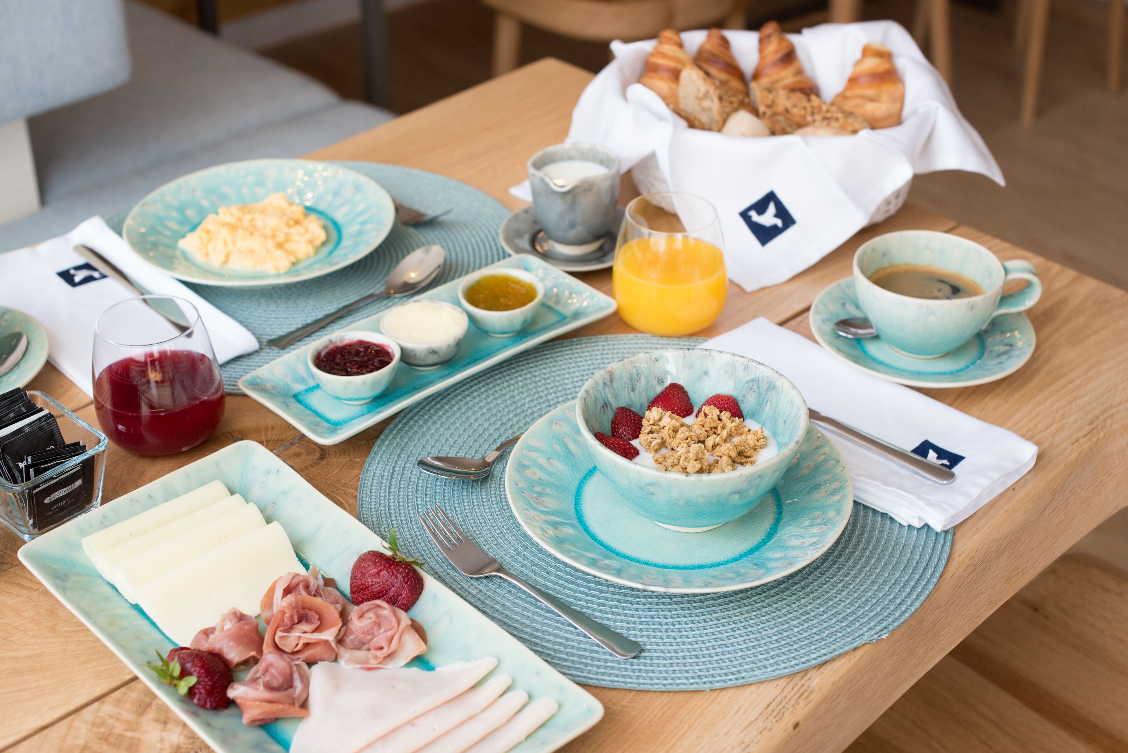 Uma mesa de pequeno-almoço com croissants, uma taça de granola com framboesas, um prato de fiambre e morangos, um copo de sumo de laranja e uma chávena de café.
