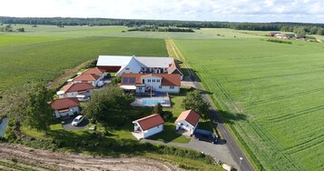 Uma casa grande com construções anexas, cercada por vastos campos verdes, uma piscina e um pequeno lago.