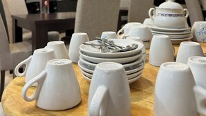 Uma área de refeições com uma mesa redonda de madeira posta com pratos, chávenas e um bule de cerâmica branca. Cadeiras rodeiam a mesa, um sofá de cor escura e um espelho na parede.