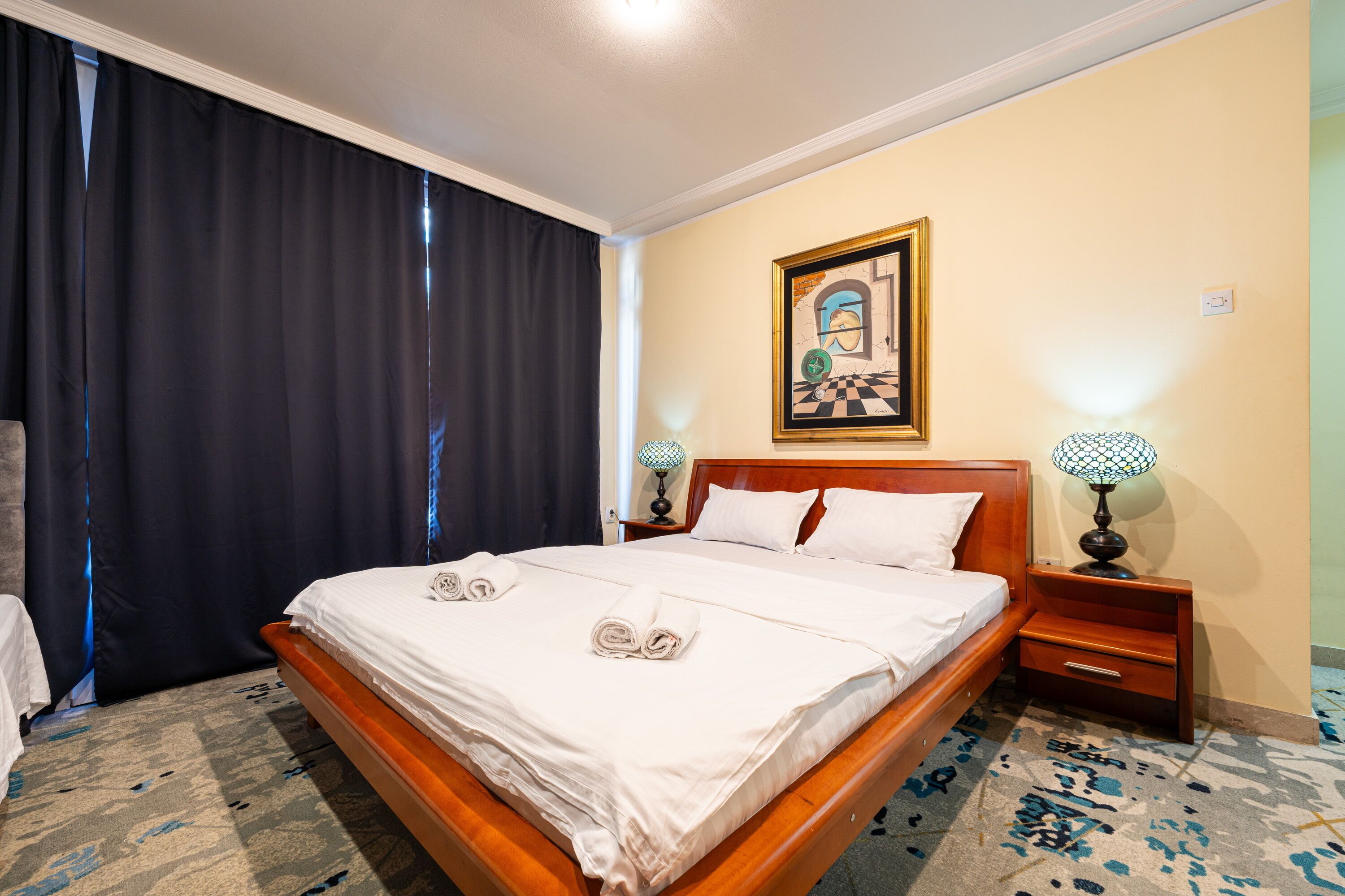 Cama bem arrumada com lençóis brancos e duas toalhas enroladas, cabeceira de madeira, mesa de cabeceira com abajur e quadro na parede.