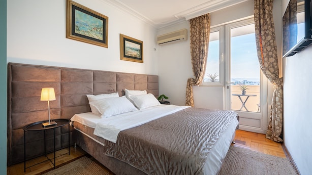 Quarto com uma cama grande, mesa de cabeceira com luminária, duas fotos emolduradas na parede e janela com cortinas.