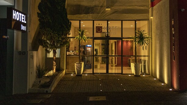 Entrada de hotel com placa indicando "HOTEL Doral", plantas em vasos grandes e um caminho pavimentado até a entrada.