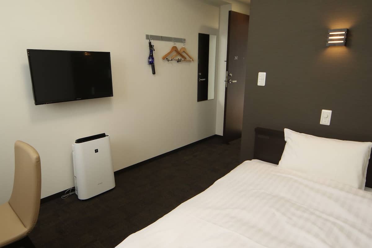 Quarto de hotel com cama, uma cadeira, televisão, cabideiro e lixeira.